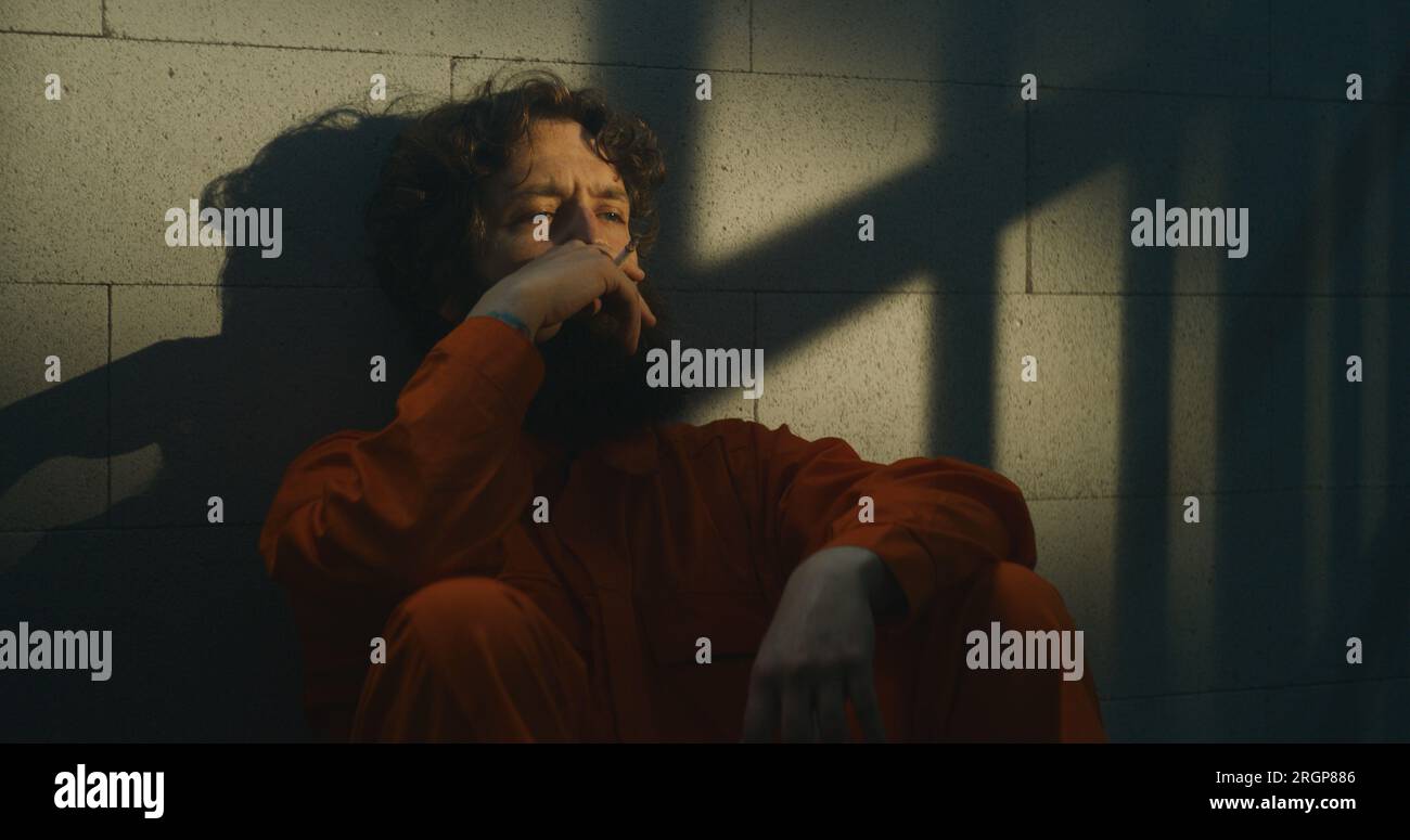 Depressed prisoner in orange uniform sits on bed, smokes cigarette in ...