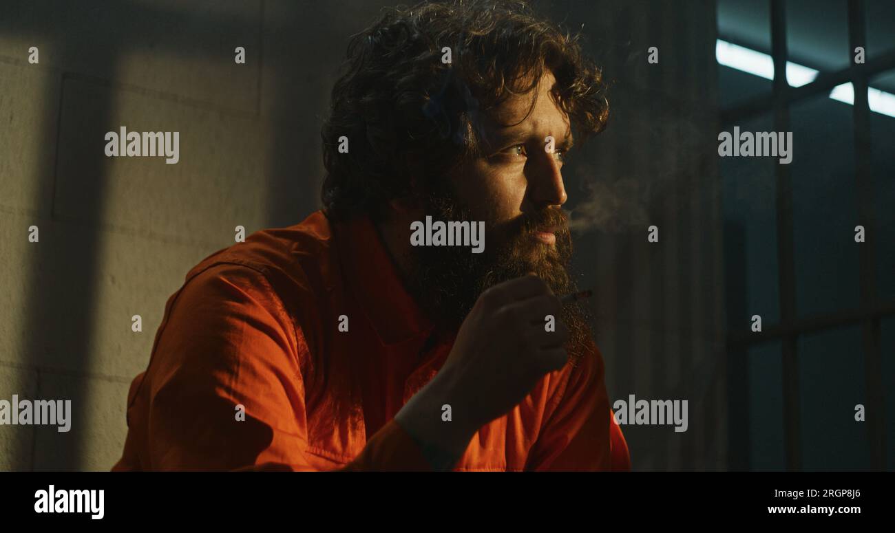 Depressed prisoner in orange uniform sits on bed in prison cell, smokes ...