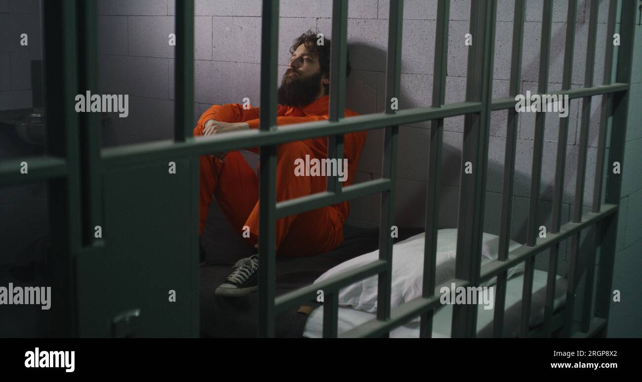 Male prisoner in orange uniform sits on bed, reads Bible, prays, looks ...