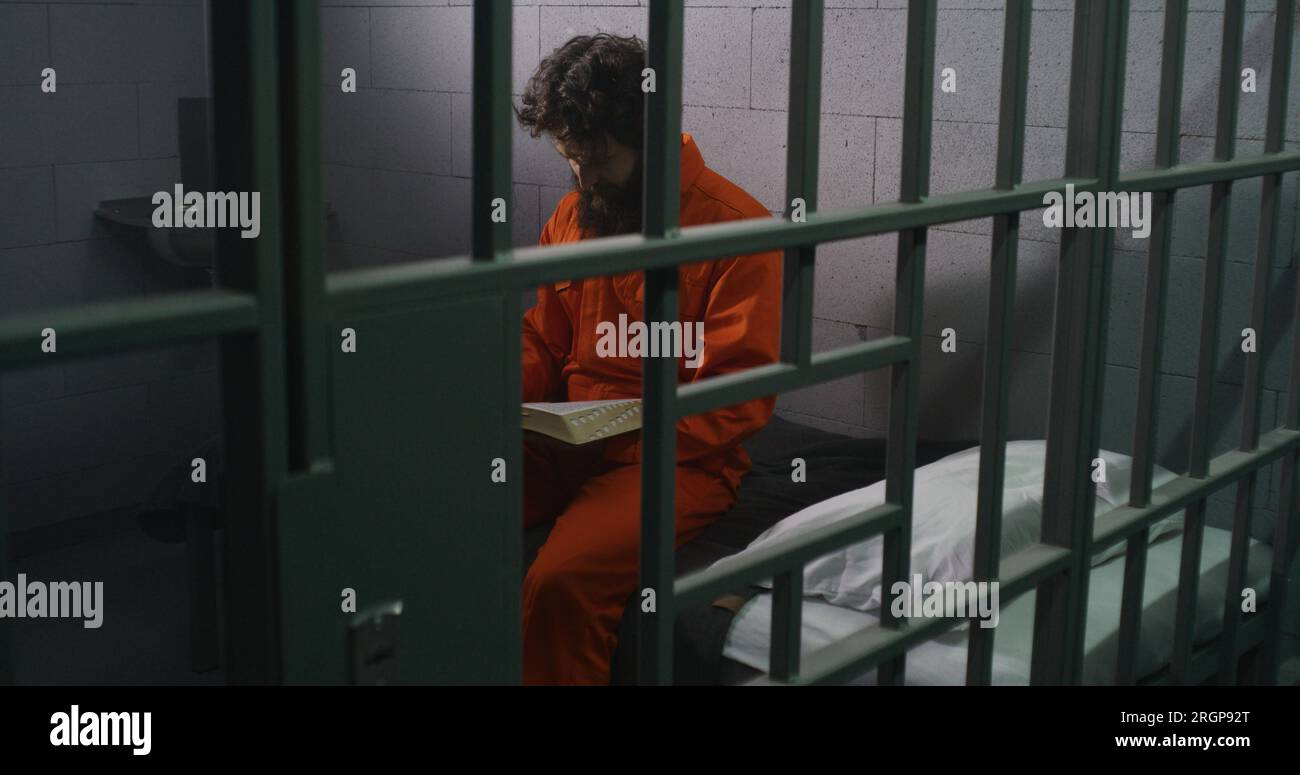 Male prisoner in orange uniform sits on bed, reads Bible, prays, looks ...