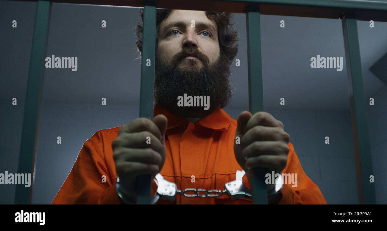 Male prisoner in orange uniform holds metal bars, stands in prison cell ...