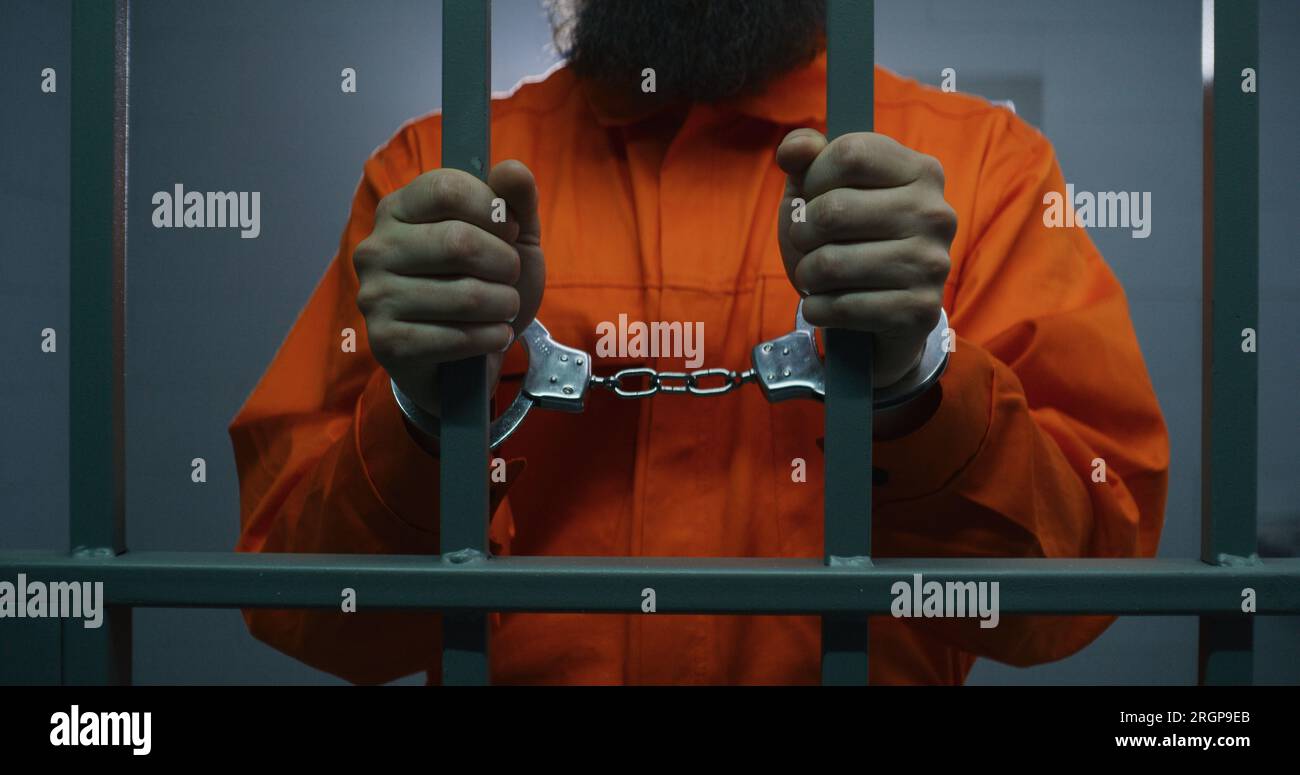 Male criminal in orange uniform and handcuffs holds metal bars, stands ...