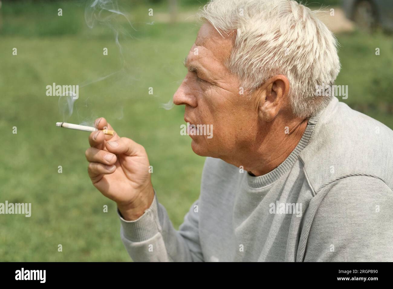 Senior man lighting up a cigarette outdoors against green nature ...