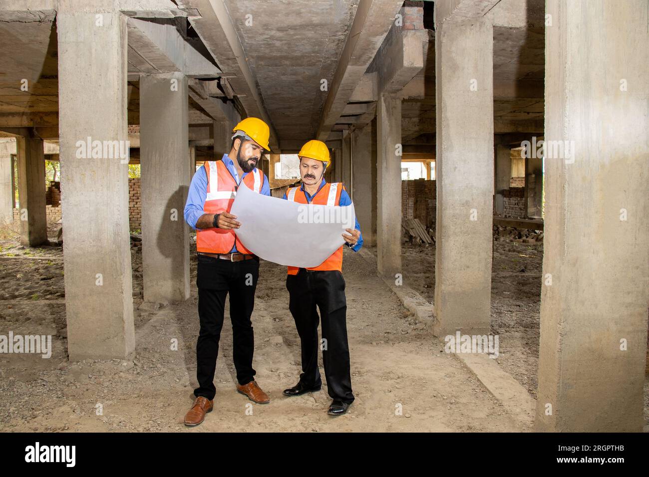 Two Indian male civil engineers or architect wearing helmet and vest holding paperwork blueprint ...