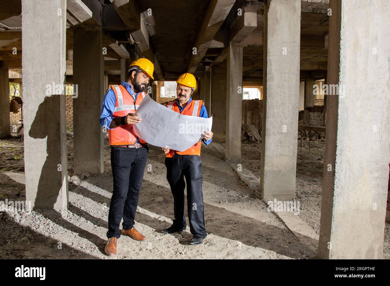 Two Indian male civil engineers or architect wearing helmet and vest holding paperwork blueprint ...