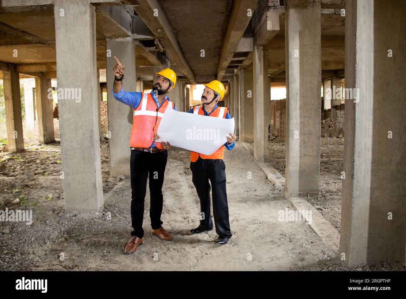 Two Indian male civil engineers or architect wearing helmet and vest holding paperwork blueprint ...