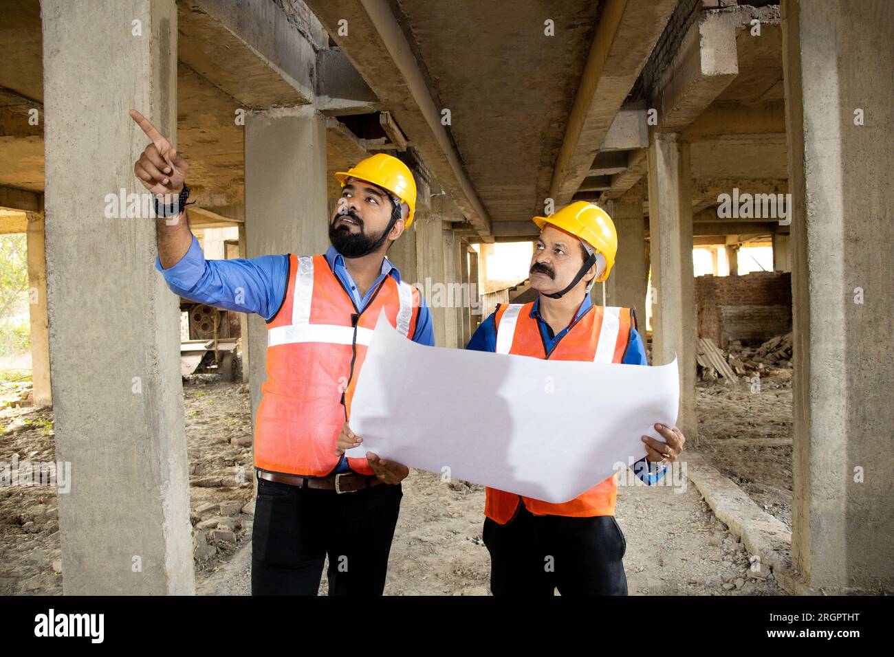 Two Indian male civil engineers or architect wearing helmet and vest holding paperwork blueprint ...