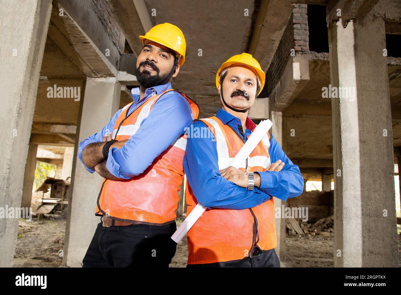 Portrait of two Indian male civil engineers or architect wearing helmet standing with cross arms ...