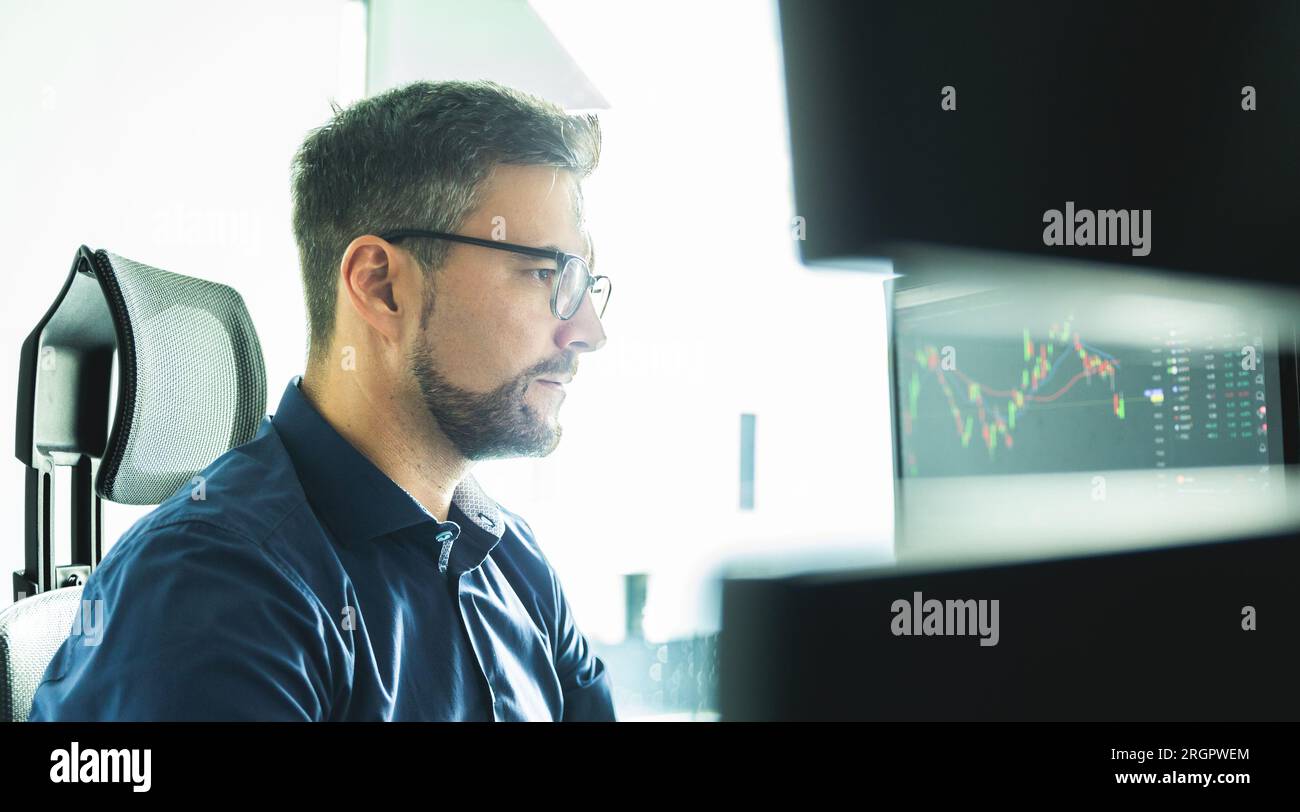 Male stock broker trading online watching charts and data analyses on multiple computer screens ...