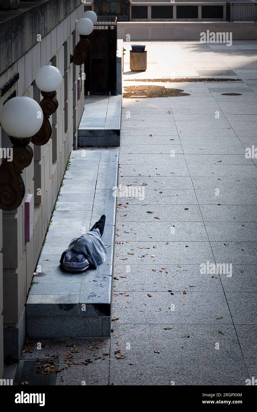A homeless person sleeps on a stone bench in front of a building near ...