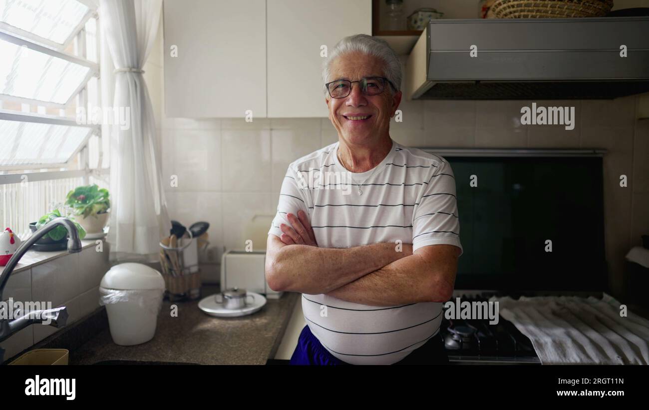 Happy senior man standing in home kitchen by window with arms crossed ...