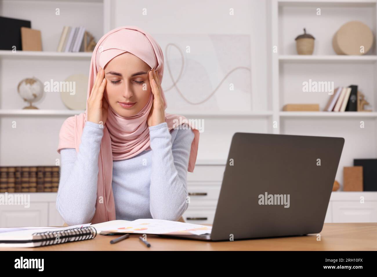 Tired Muslim woman in hijab working near laptop at wooden table in room ...