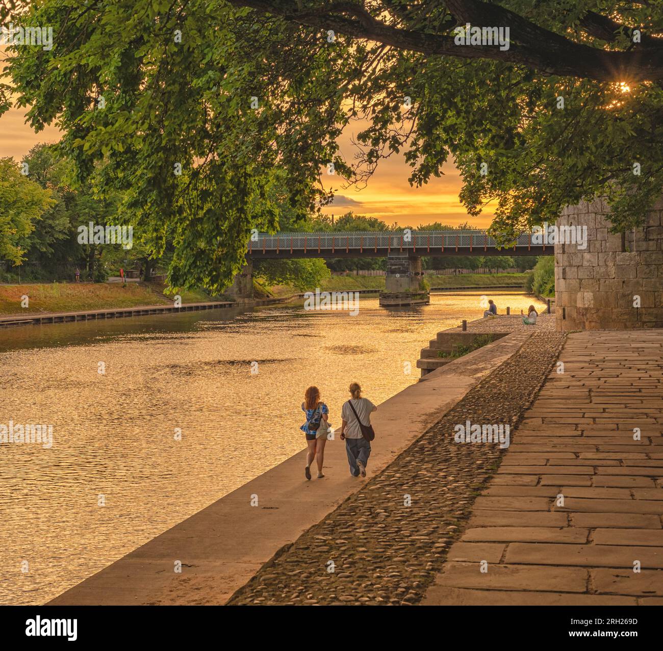 A couple walk down a riverside path at sunset towards an ancient ...