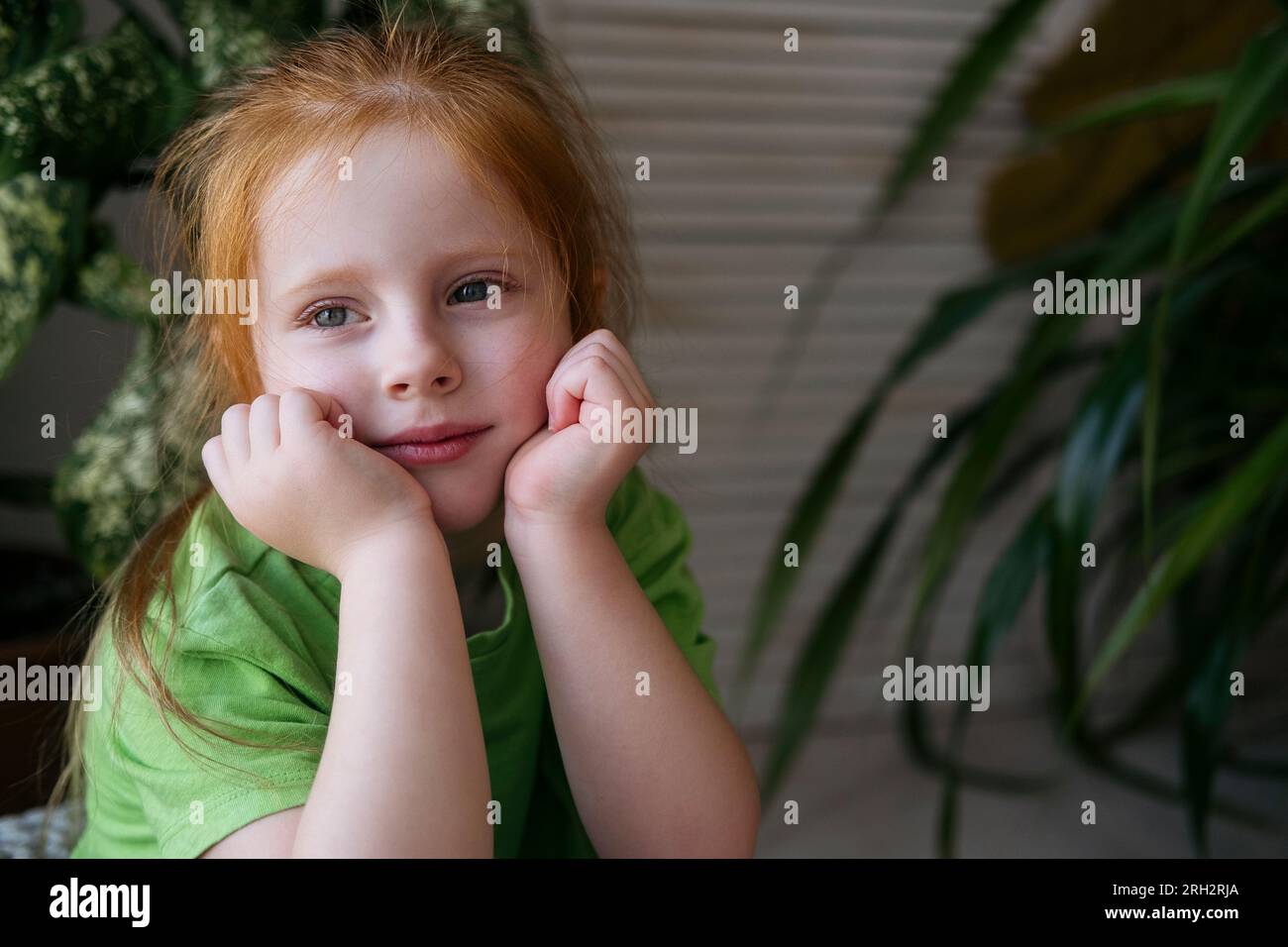 A red-haired little girl in a green T-shirt sits near the window at home and smiles Stock Photo ...