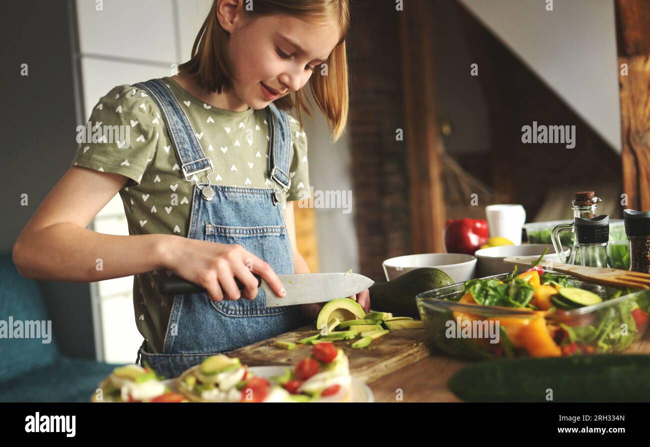 Girl child cuts avicado with knife at kitchen. Pretty female kid with ...