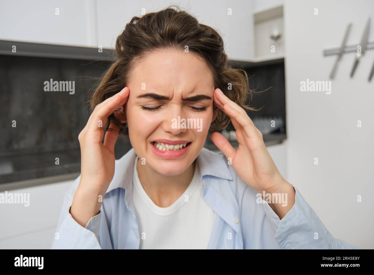 Portrait of woman with headache, sits in kitchen, touches head and ...