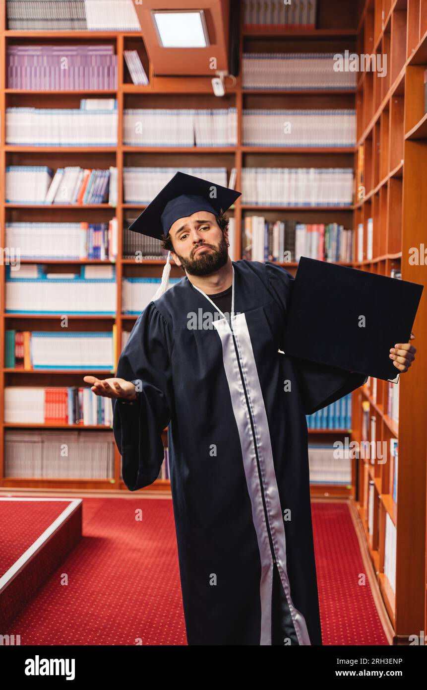 Young graduate wearing his cap and gown looking sad while holding his ...