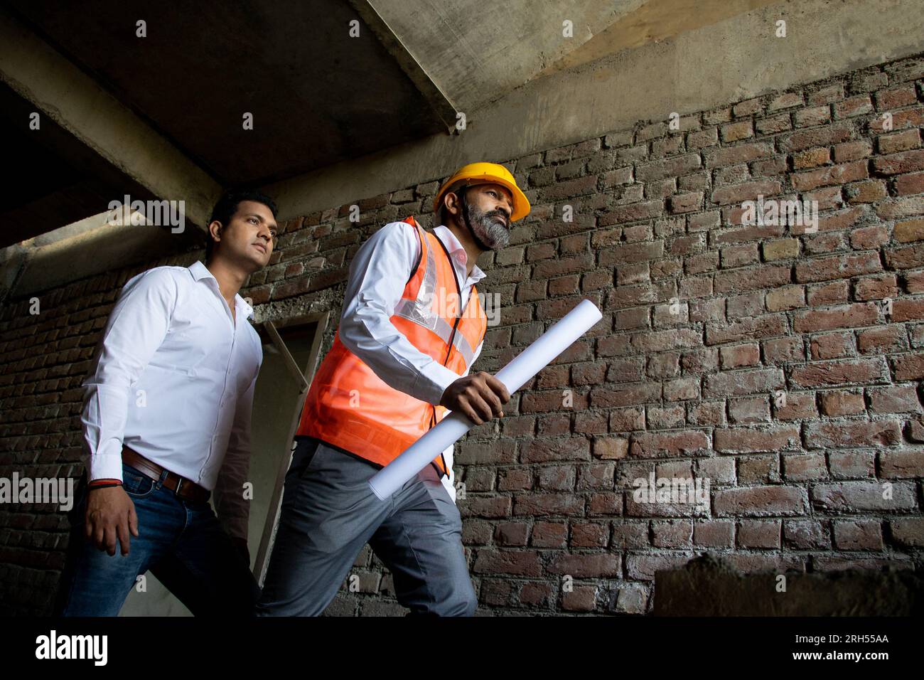 Two Indian male civil engineers or architect wearing helmet and vest holding paperwork blueprint ...
