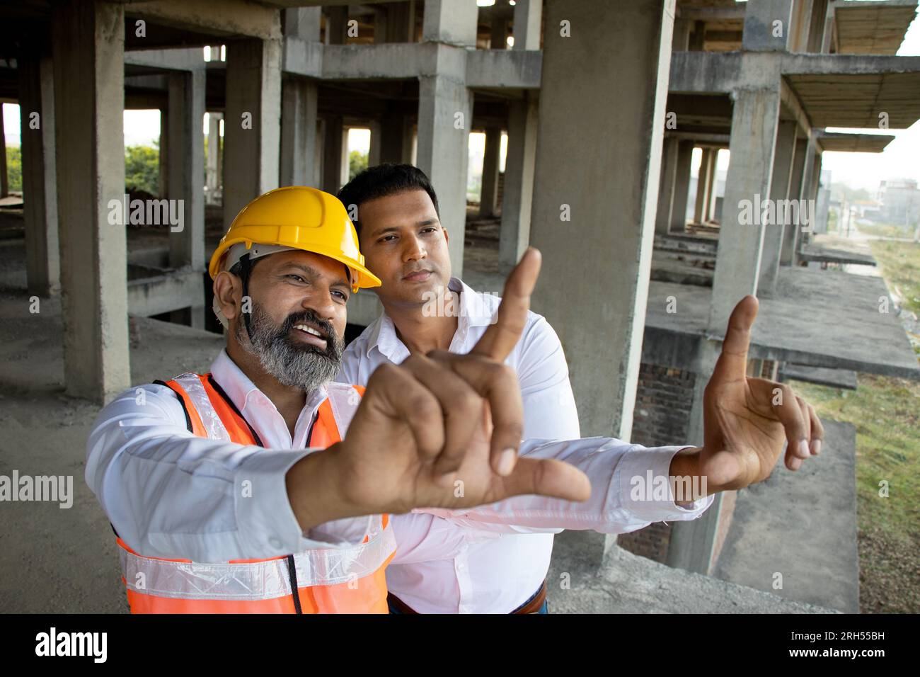 Two Indian male civil engineers or architect wearing helmet and vest holding paperwork blueprint ...
