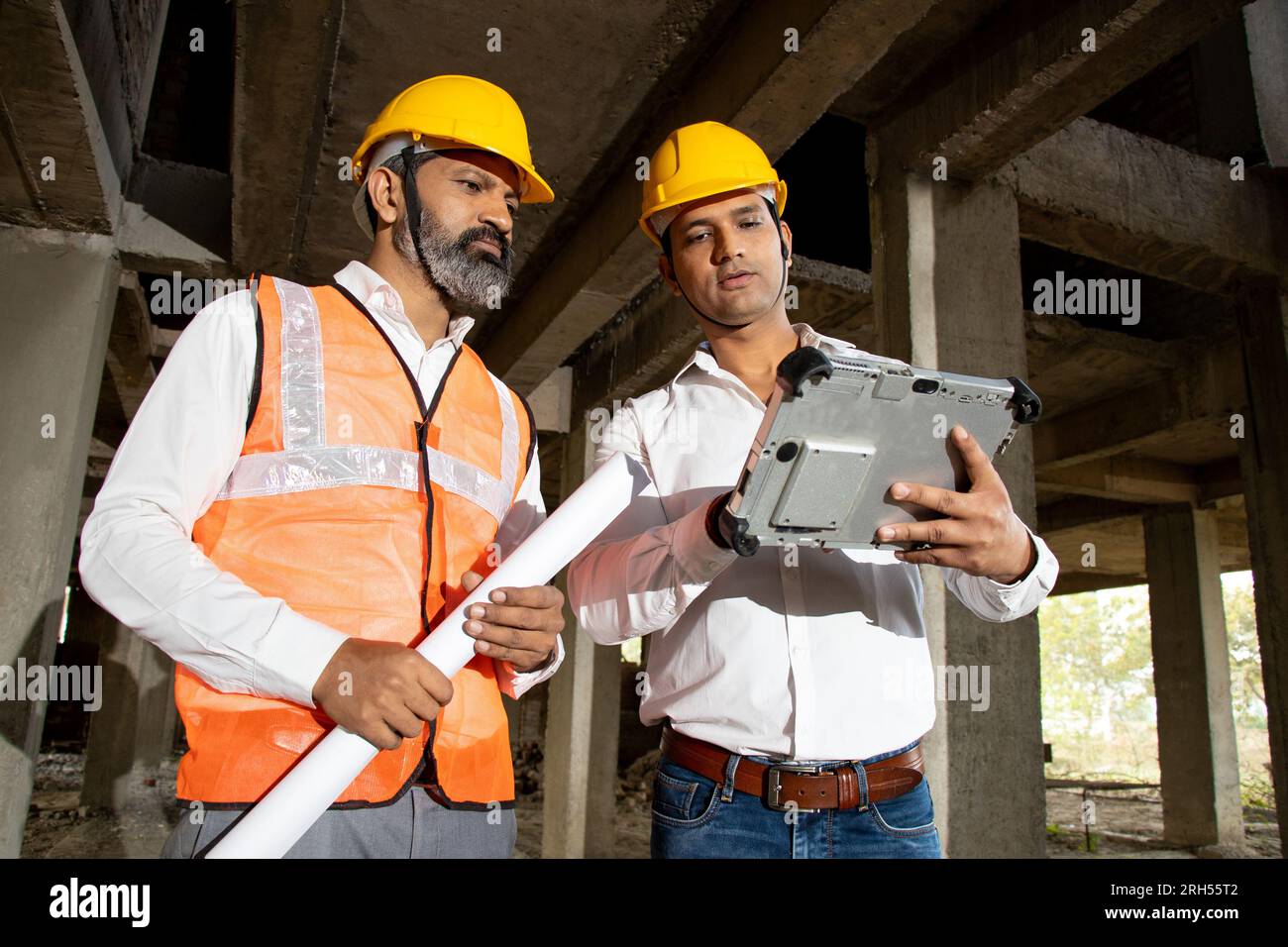 Two Indian male civil engineers or architect wearing helmet and vest holding digital tablet ...