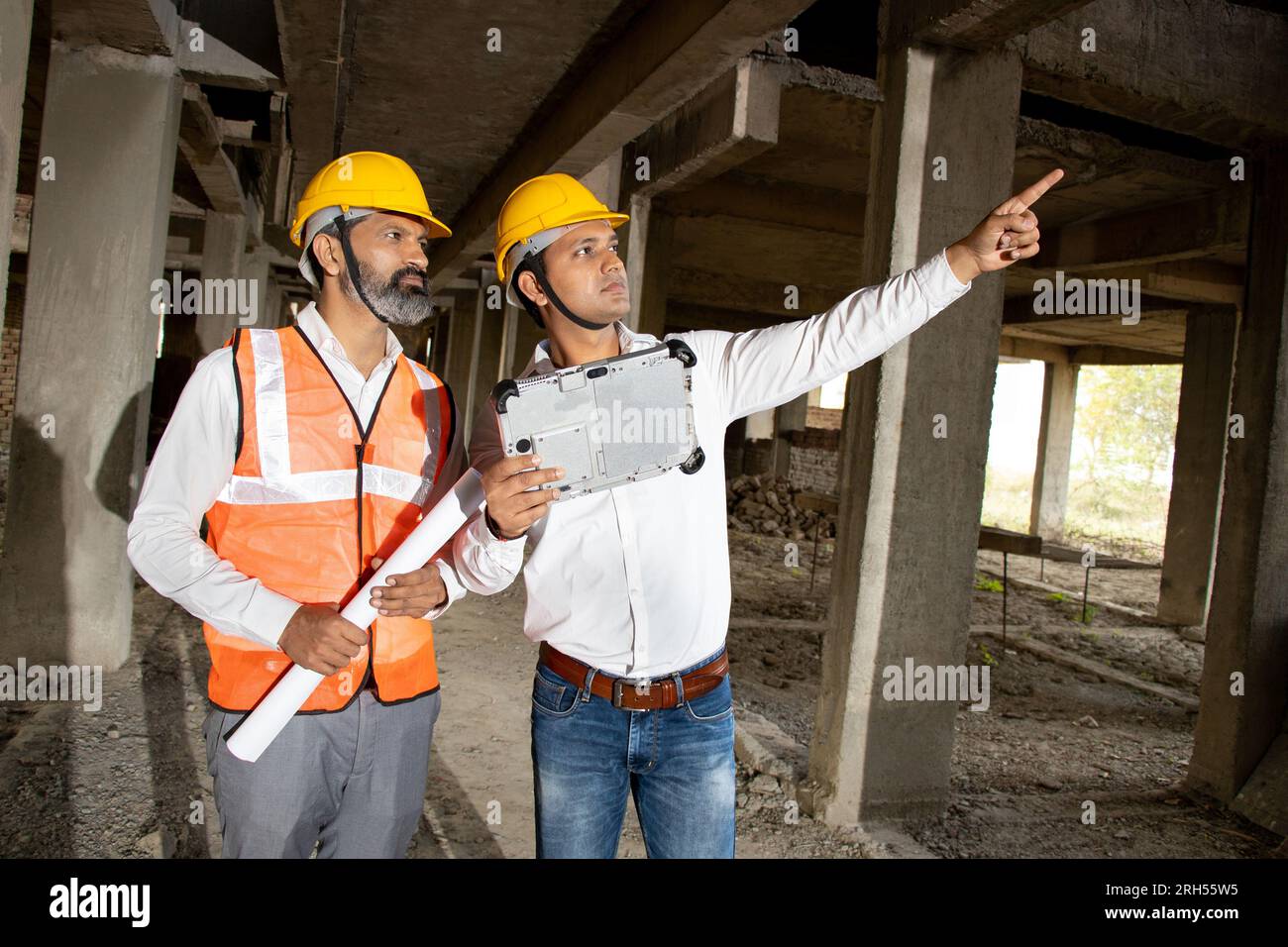 Two Indian male civil engineers or architect wearing helmet and vest holding paperwork blueprint ...