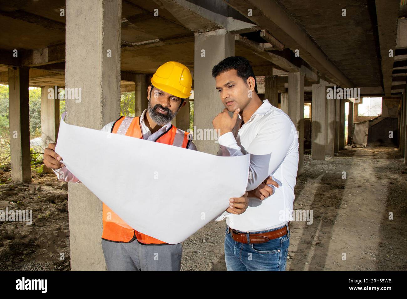 Two Indian male civil engineer or architect wearing helmet and vest holding paperwork blueprint ...