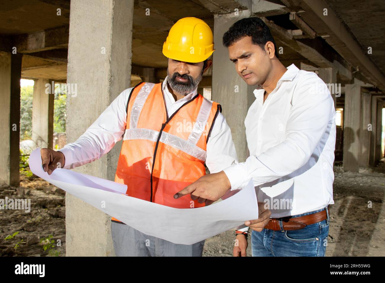 Two Indian male civil engineer or architect wearing helmet and vest holding paperwork blueprint ...