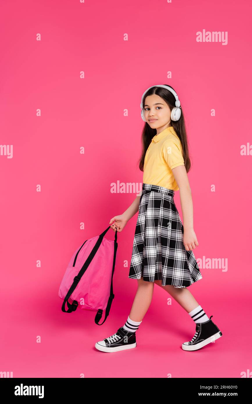 brunette schoolgirl in wireless headphones holding backpack, walking on ...