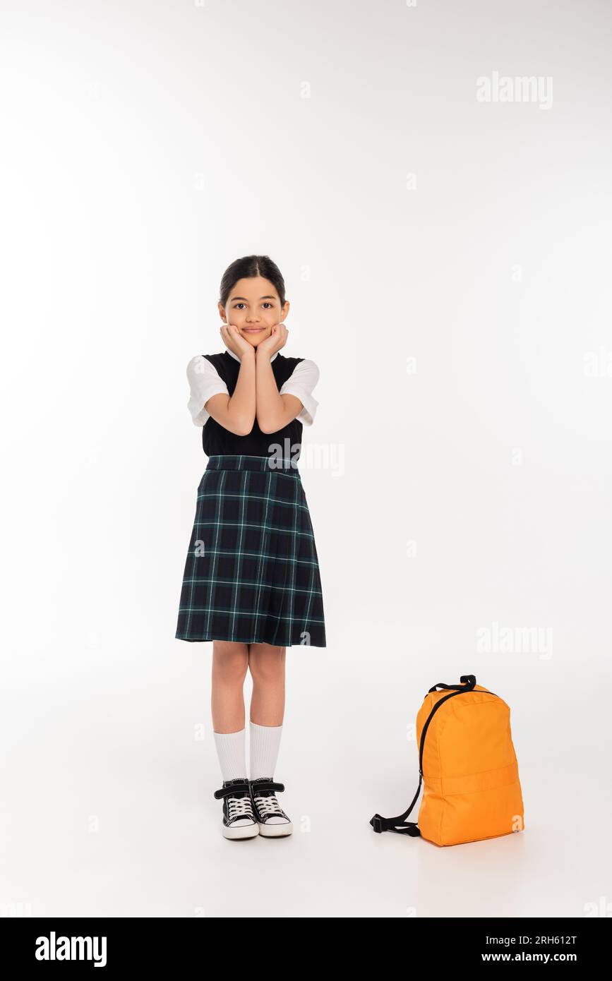 happy girl in school uniform standing near backpack on white background ...