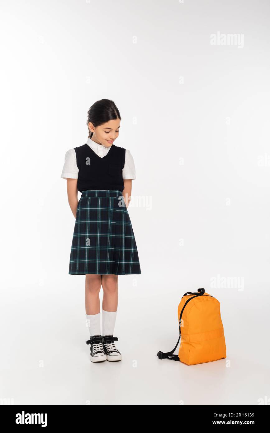 happy girl in school uniform standing and looking at backpack on white ...