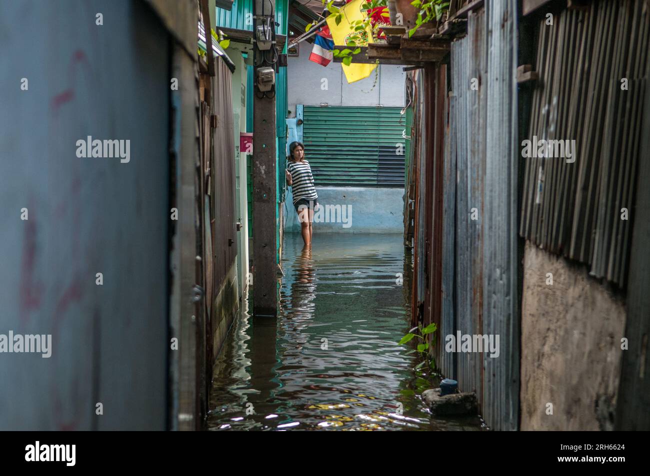A young Thai woman stands in floodwater, down a poverty-stricken alley ...