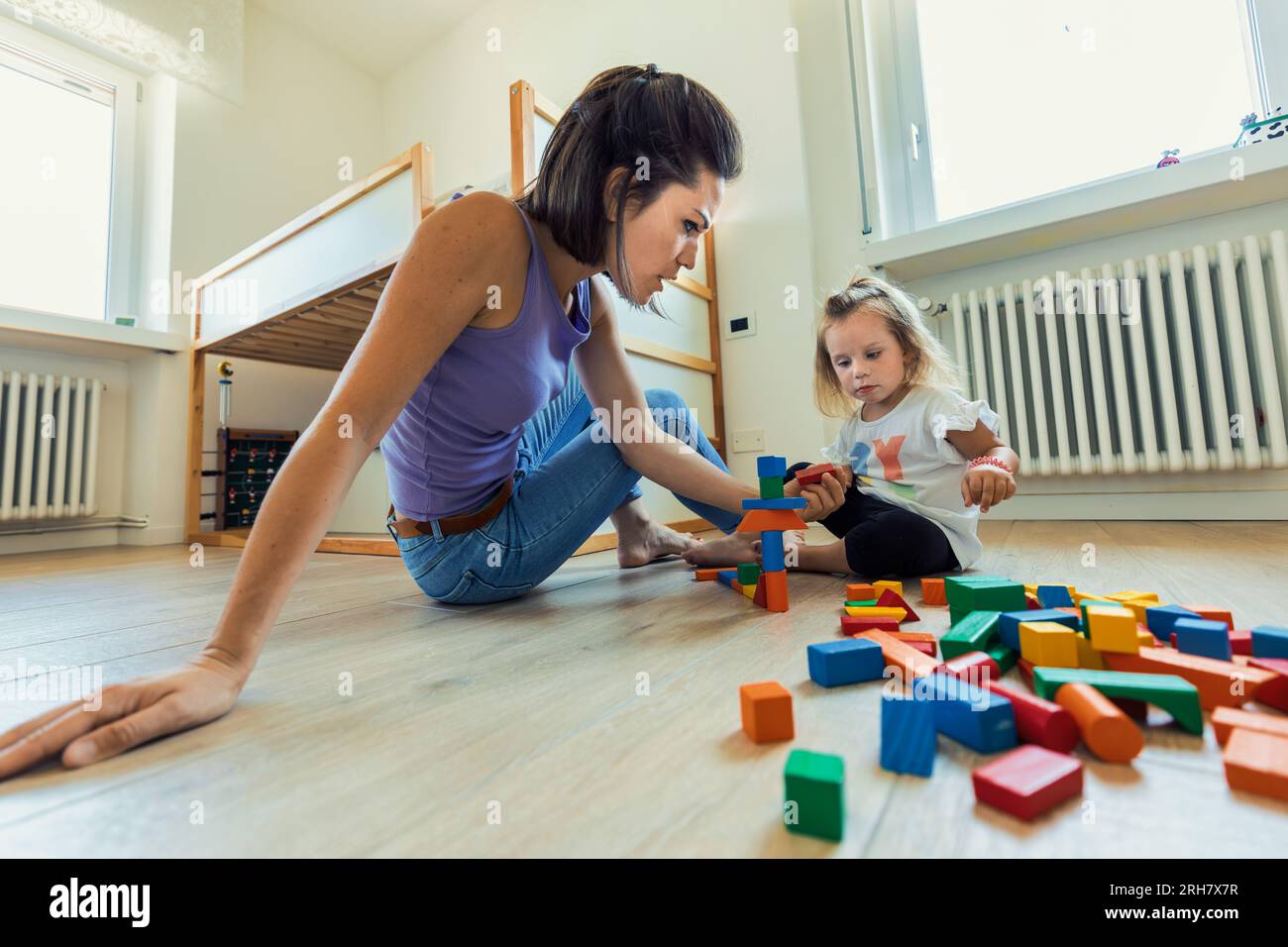 Mother and daughter engage in the playroom with wooden blocks, cups, developing skills, enjoying ...