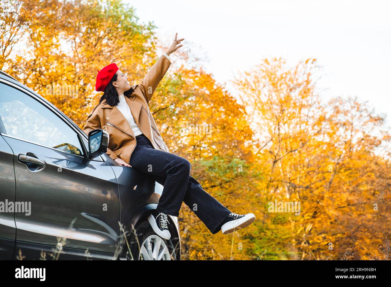Stylish woman in a cute outfit sits on the hood of an automobile with ...