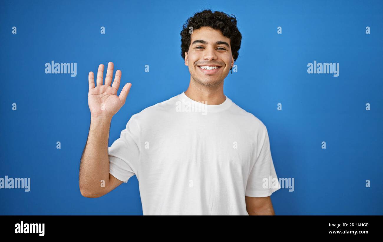 Young latin man saying hello with hand smiling over isolated blue ...