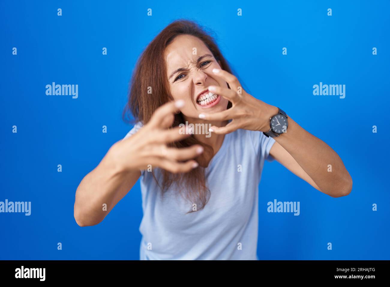 Brunette woman standing over blue background shouting frustrated with ...
