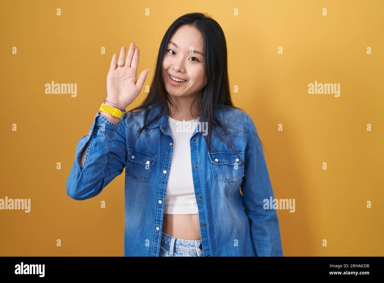 Young asian woman standing over yellow background waiving saying hello ...