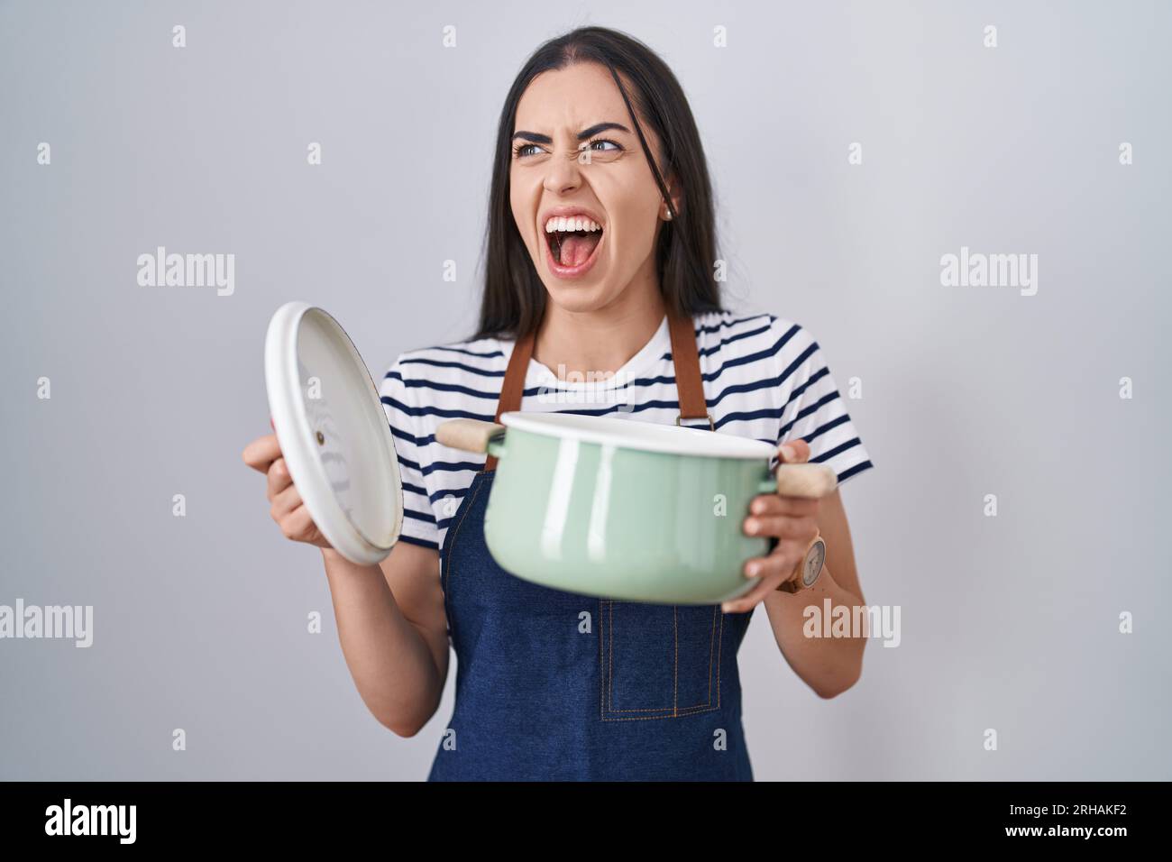 Young brunette woman wearing apron holding cooking pot angry and mad ...