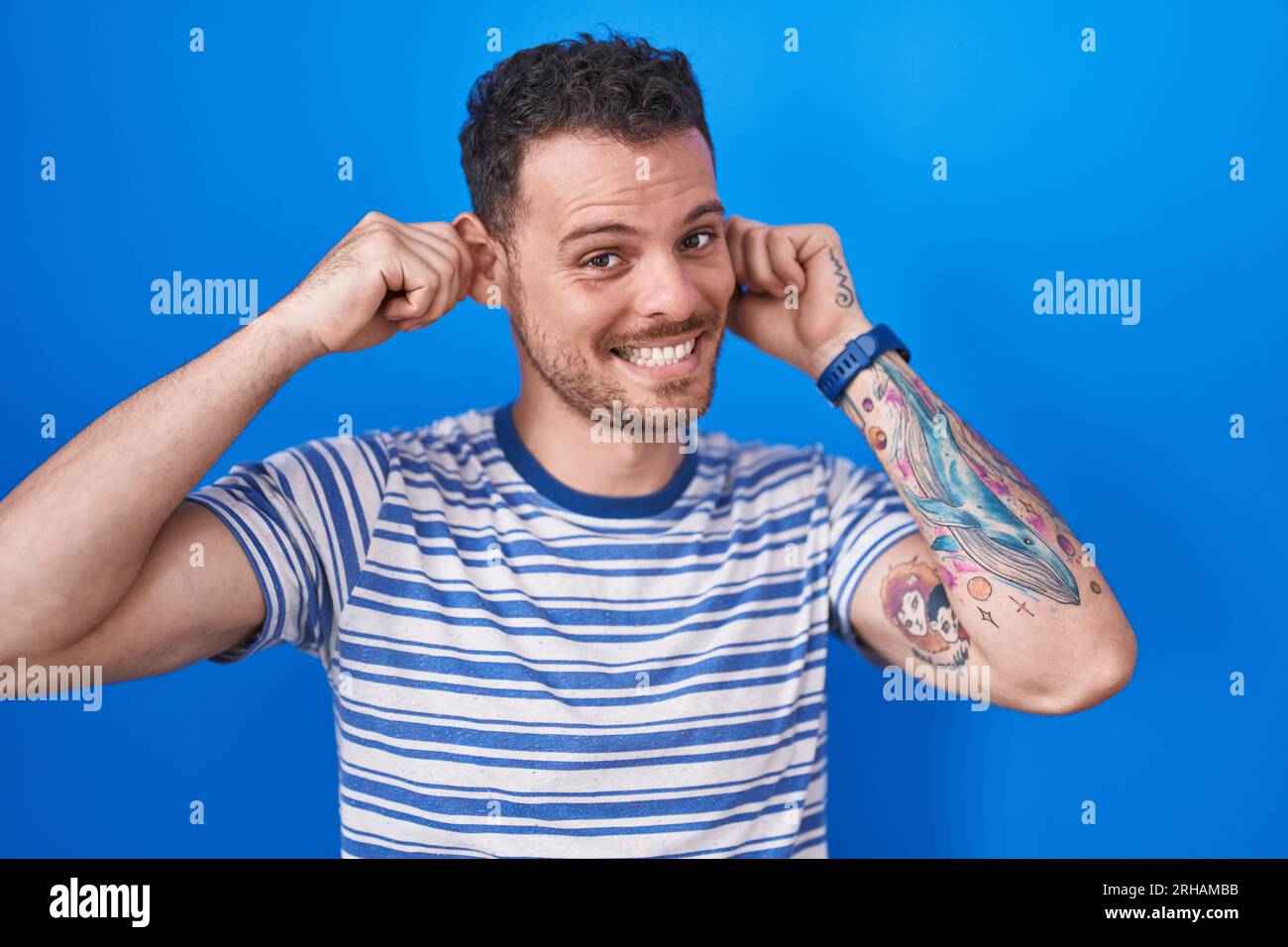 Young hispanic man standing over blue background smiling pulling ears ...