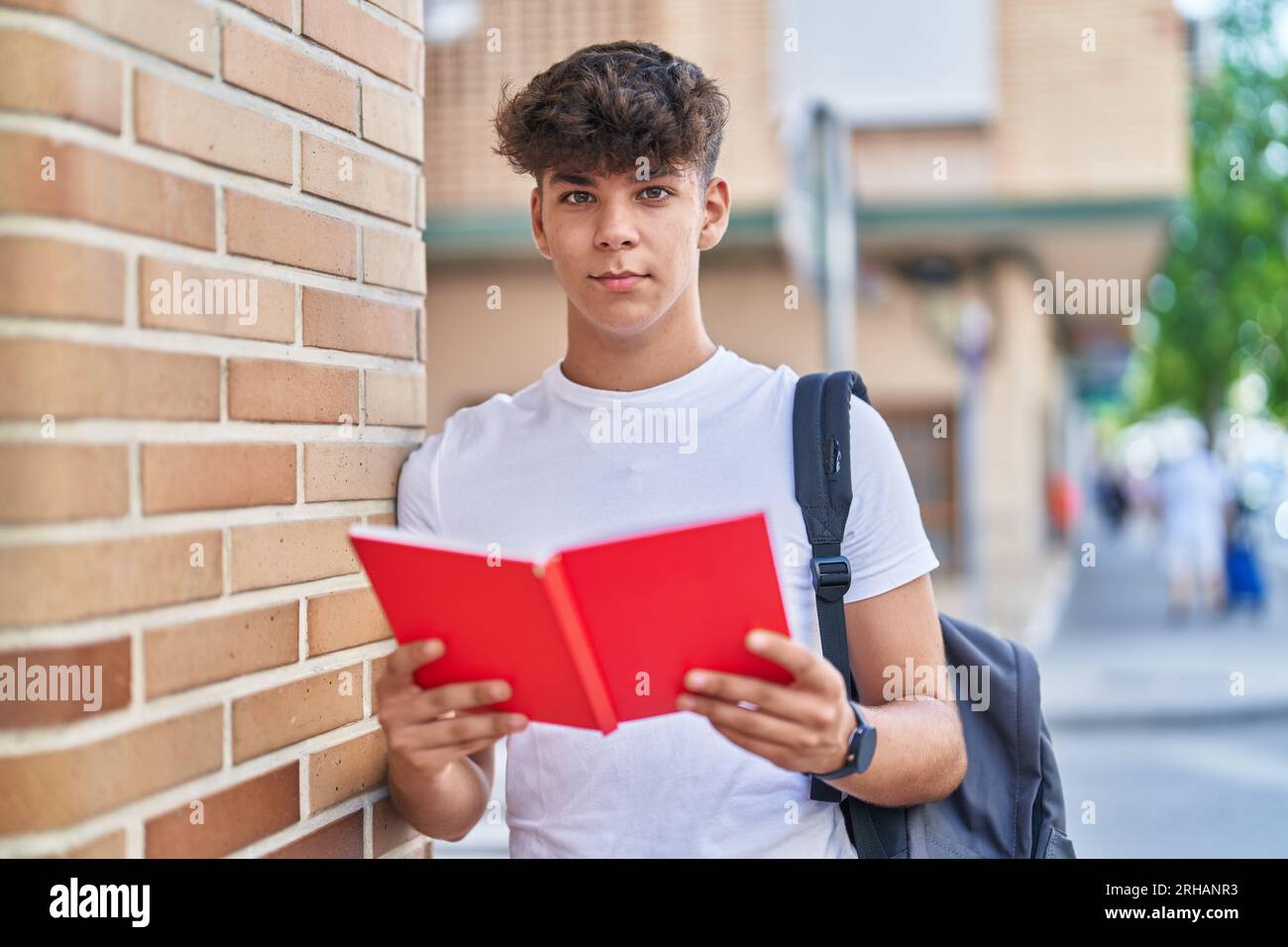 Young hispanic teenager student wearing backpack reading book at street ...