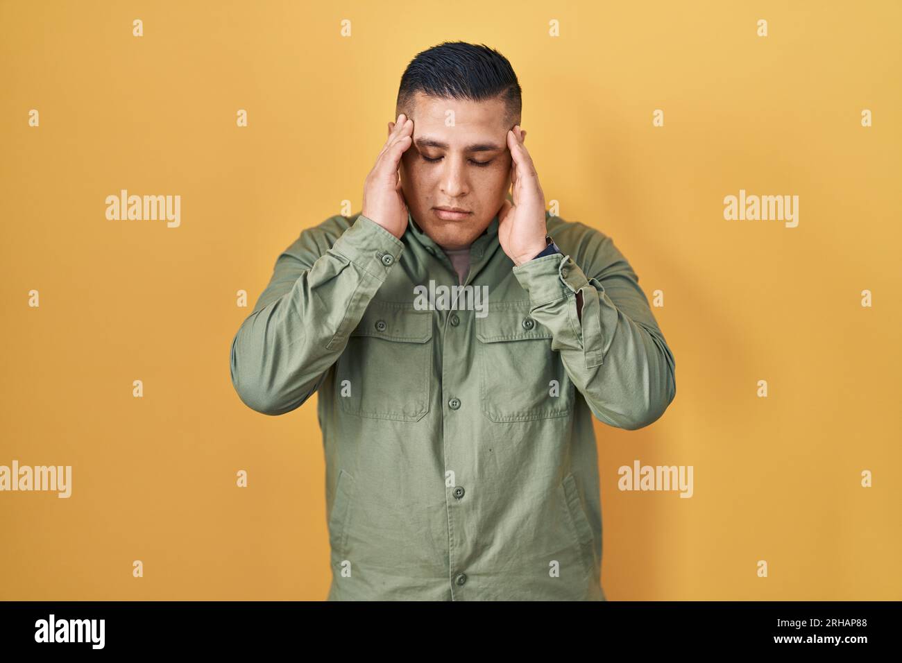 Hispanic young man standing over yellow background with hand on head ...