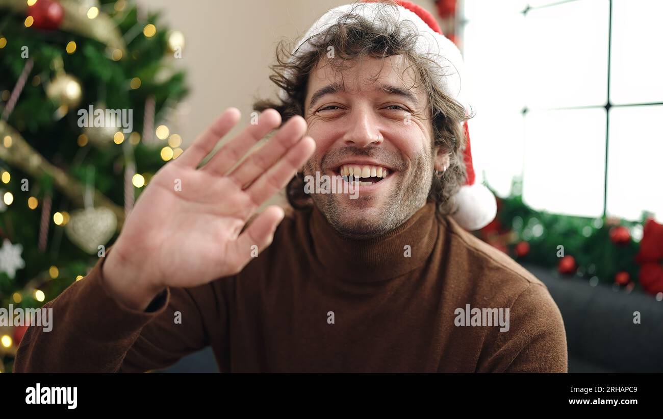 Young hispanic man saying hello with hand sitting on sofa by christmas ...