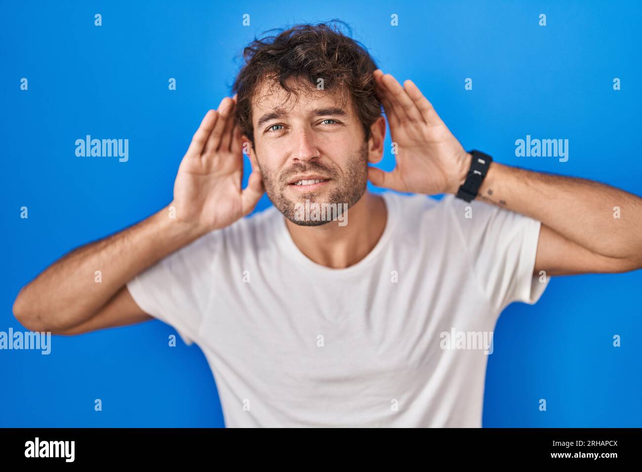 Hispanic young man standing over blue background trying to hear both ...