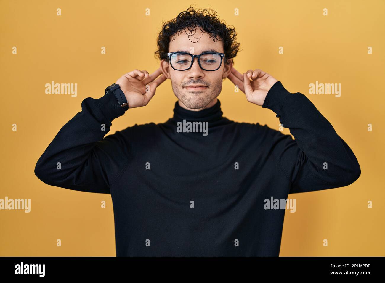 Hispanic man standing over yellow background smiling pulling ears with ...