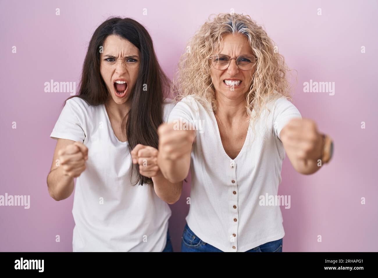Mother and daughter standing together over pink background angry and ...