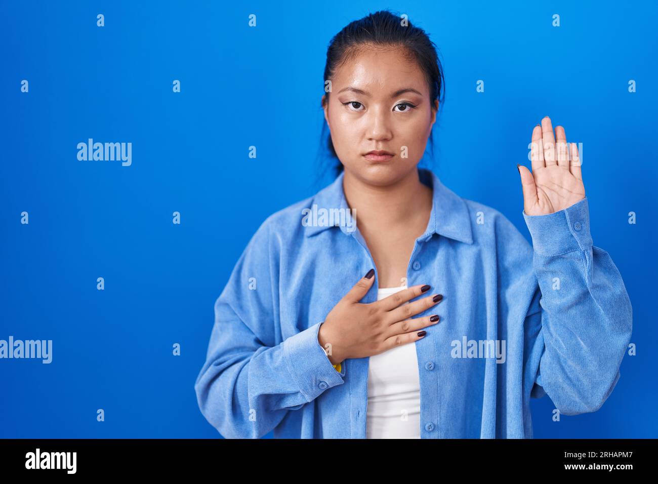 Asian young woman standing over blue background swearing with hand on ...