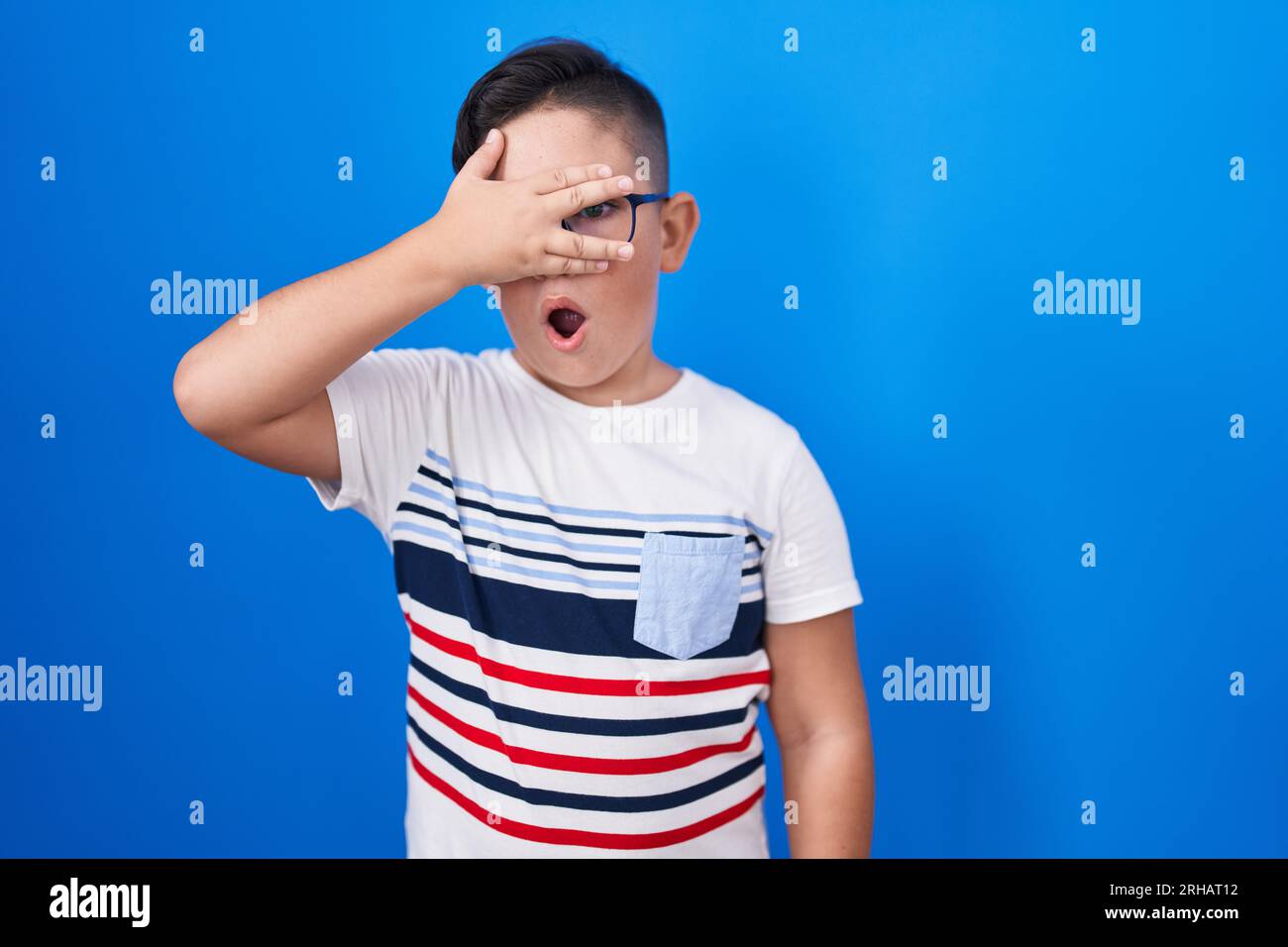 Young hispanic kid standing over blue background peeking in shock ...