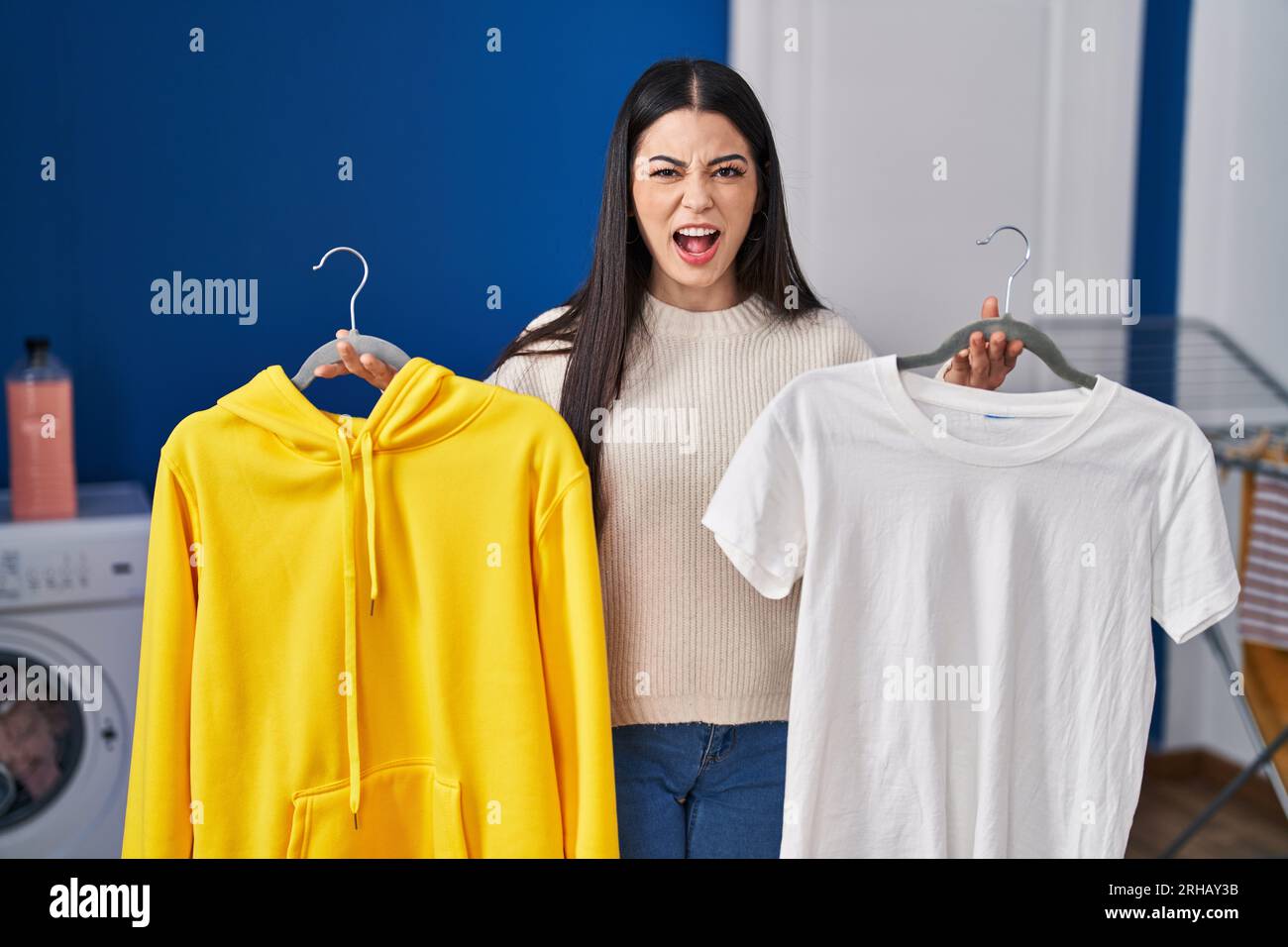 Young woman hanging clothes on racks angry and mad screaming frustrated ...