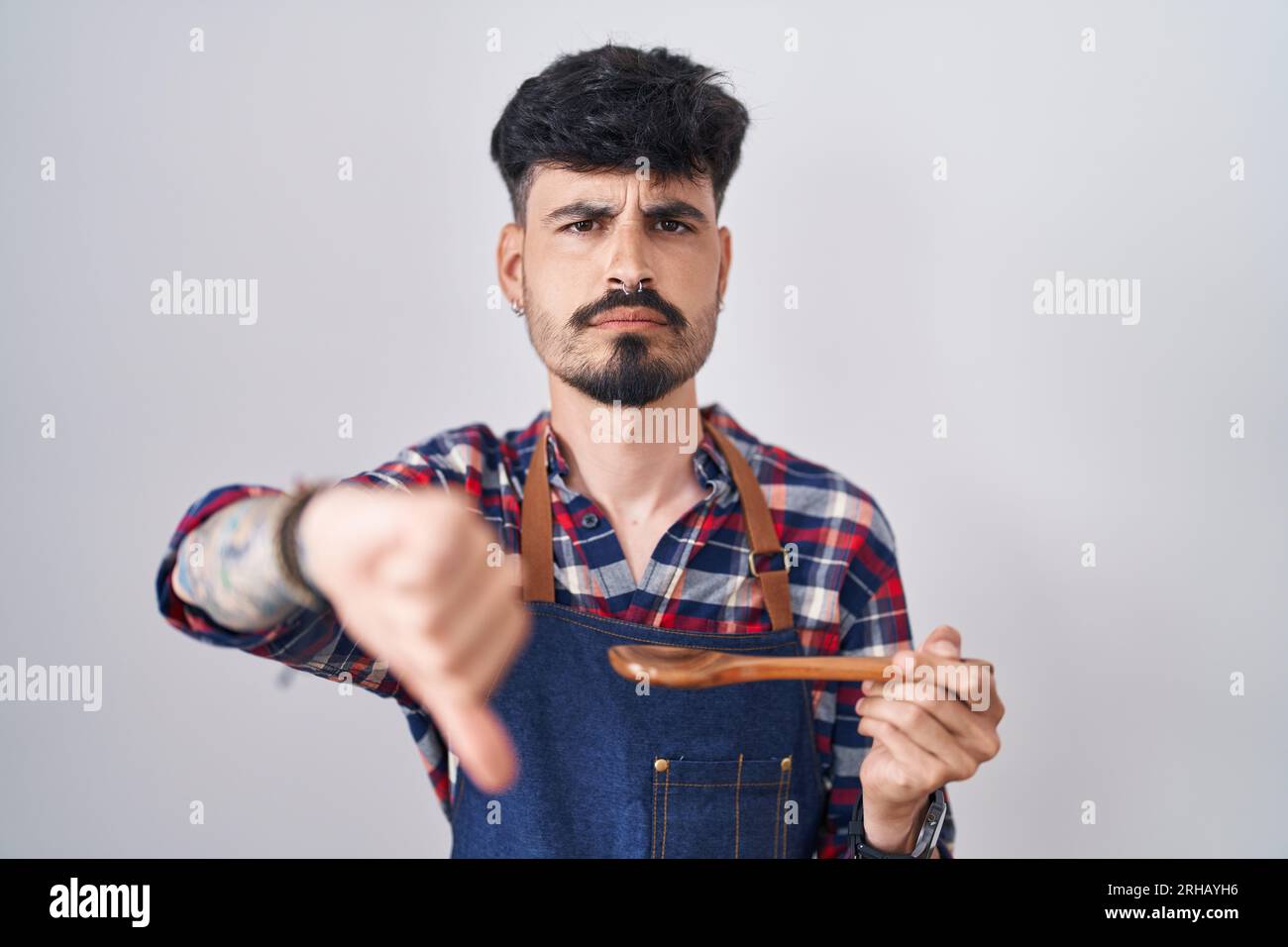 Young hispanic man with beard wearing apron tasting food holding wooden ...