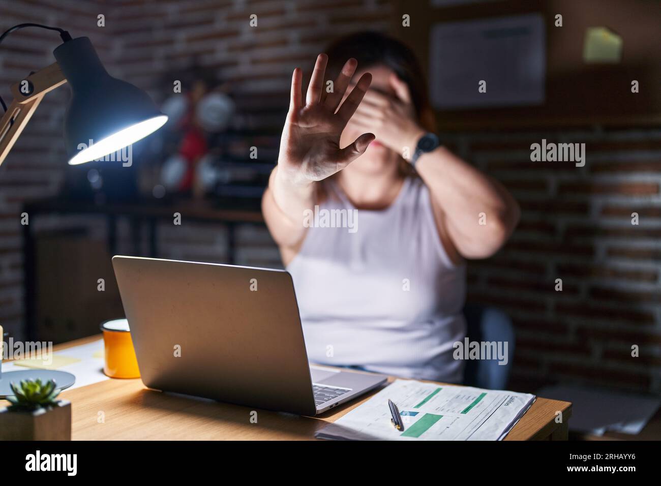 Brunette woman working at the office at night covering eyes with hands ...