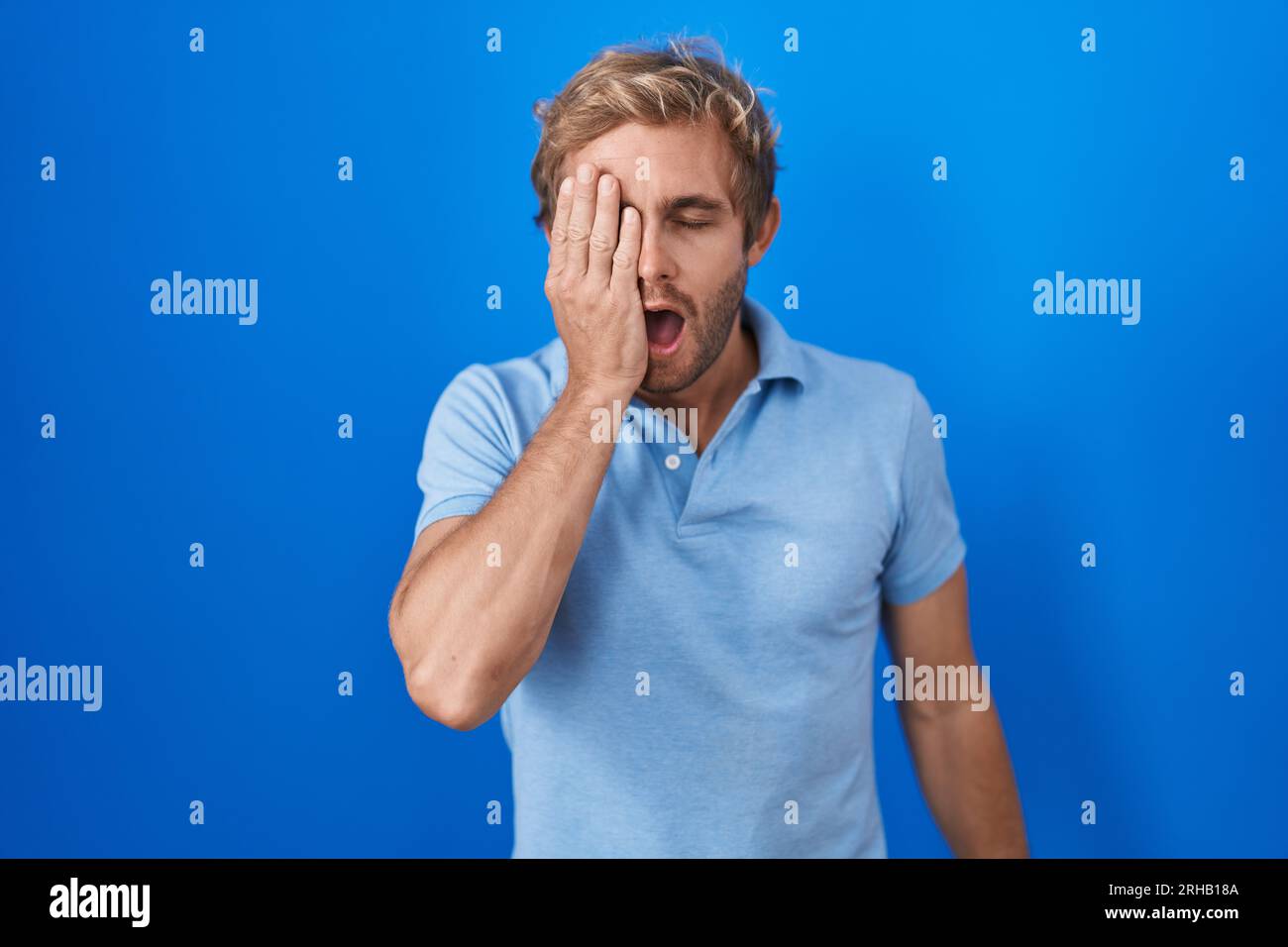 Caucasian man standing over blue background yawning tired covering half ...